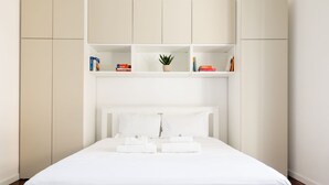 Cama bem arrumada com lençóis brancos e duas toalhas, um pequeno vaso de planta em uma prateleira acima e alguns livros na prateleira.