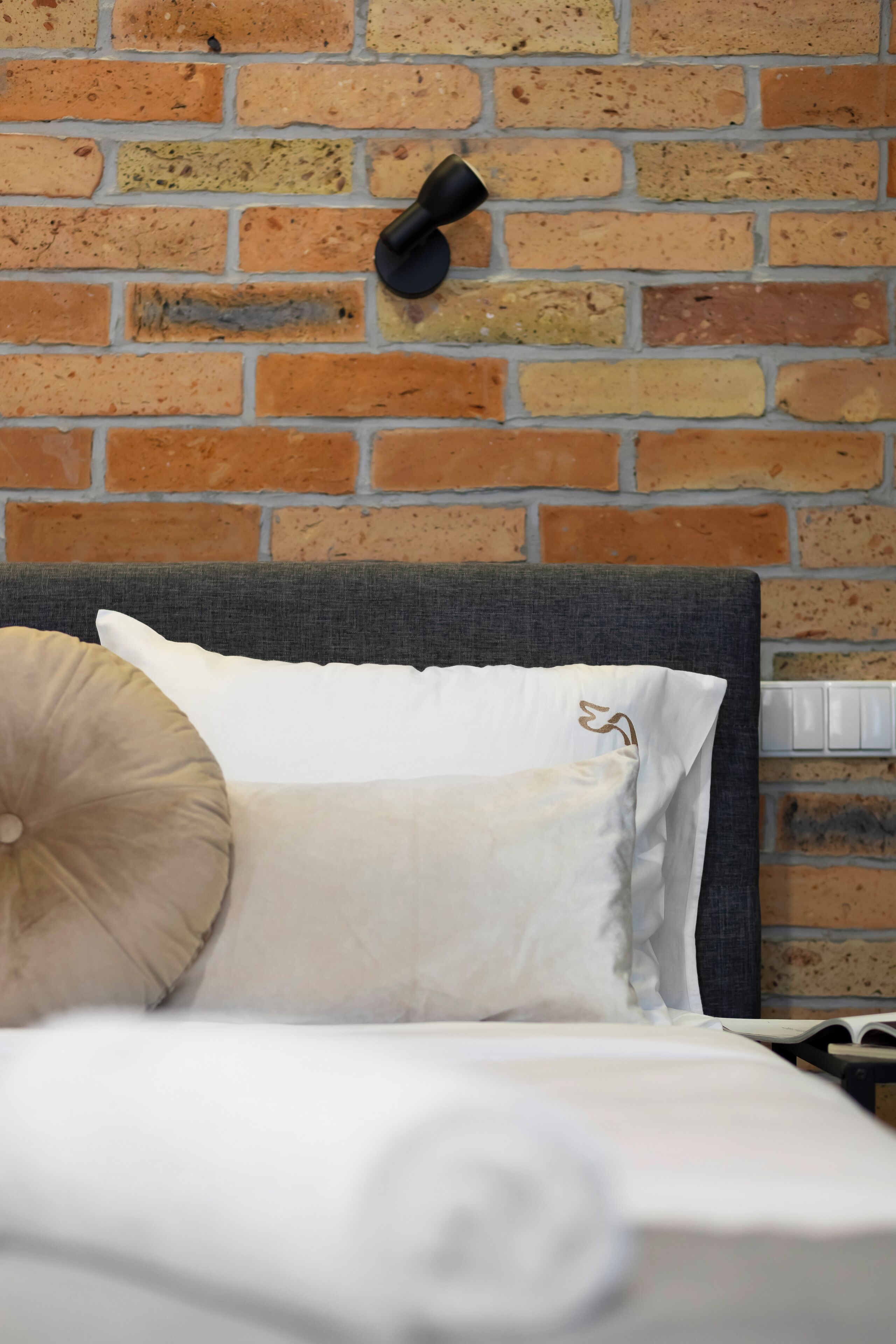 Cama com roupa de cama branca e travesseiro bege, cabeceira cinza e parede de tijolos com luminária preta fixada.