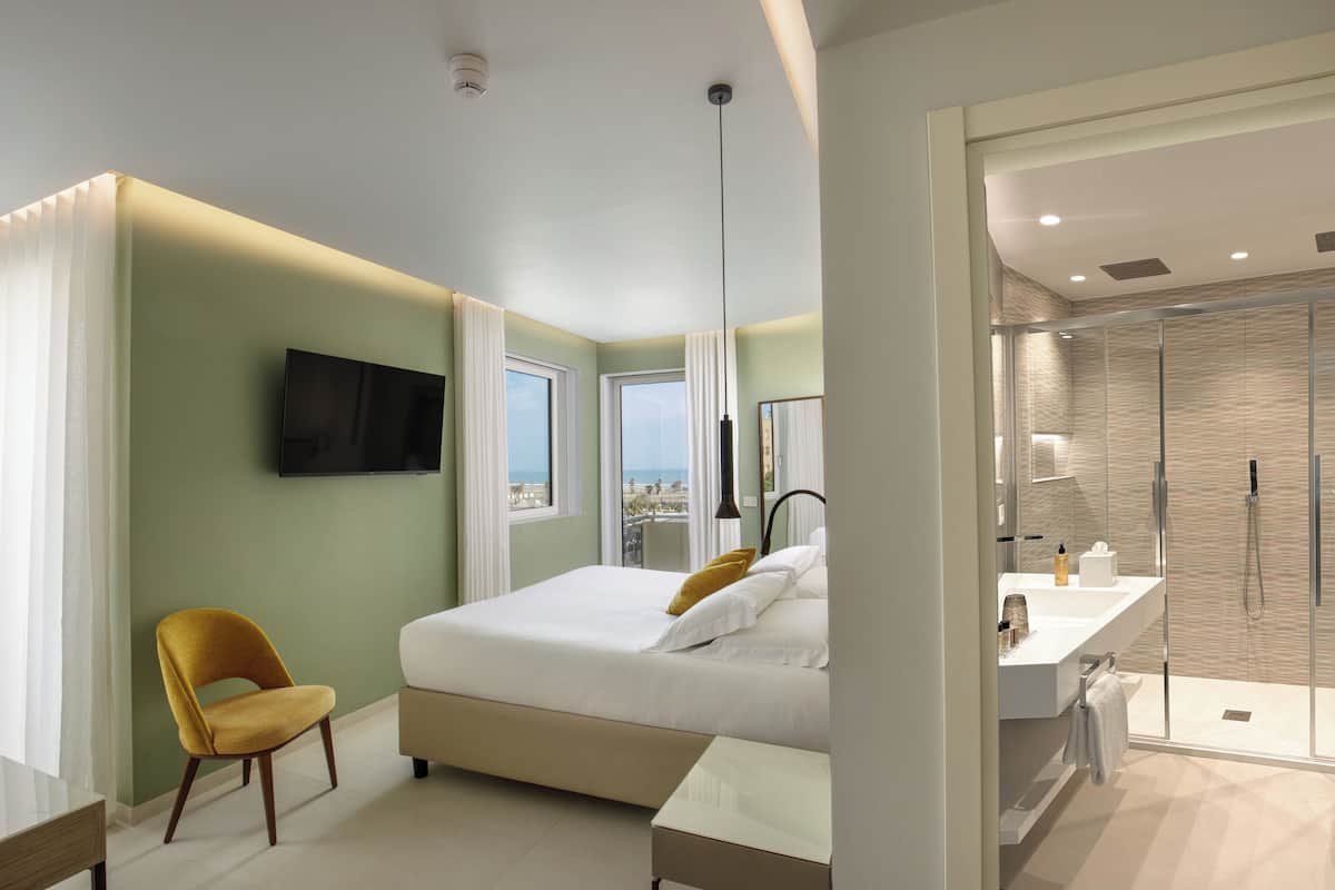 Quarto moderno com cama, poltrona, televisão, banheiro com chuveiro e vista para o mar.