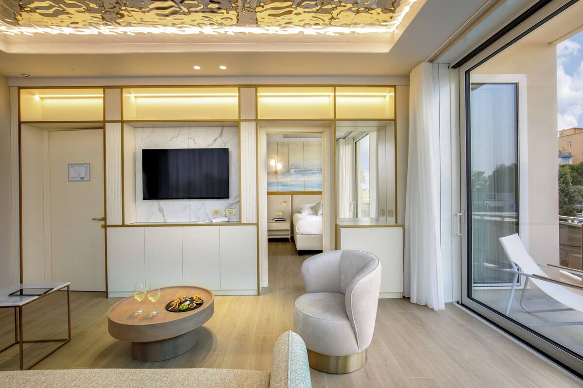 Quarto de hotel moderno com televisão grande, área de refeições, sofá e varanda com vista.