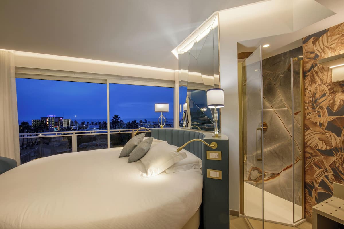 Quarto amplo com uma cama grande, chuveiro com box de vidro e vista para a cidade.