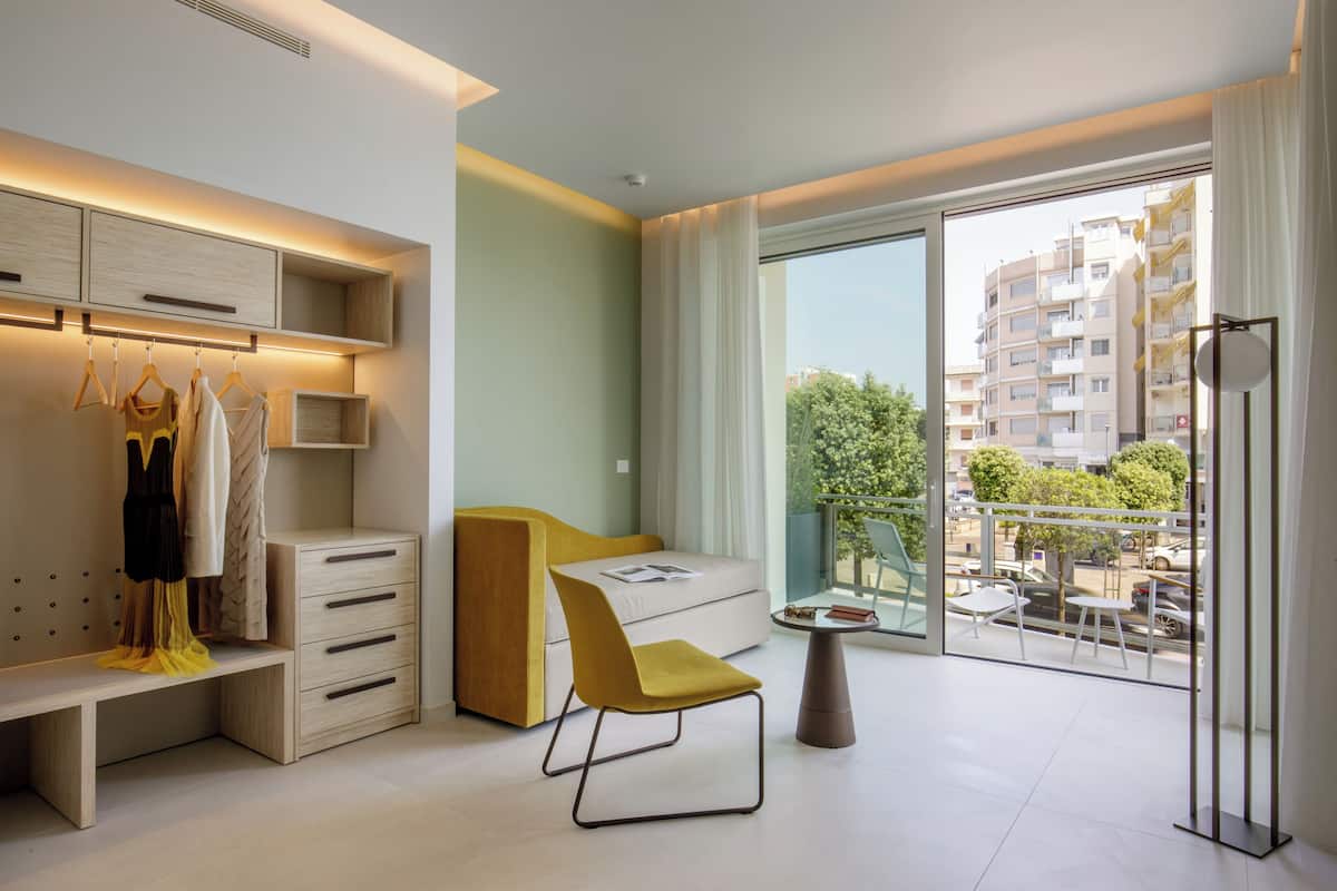 Quarto de hotel moderno com sofá, uma cadeira, uma mesinha e uma varanda com vista para a cidade.