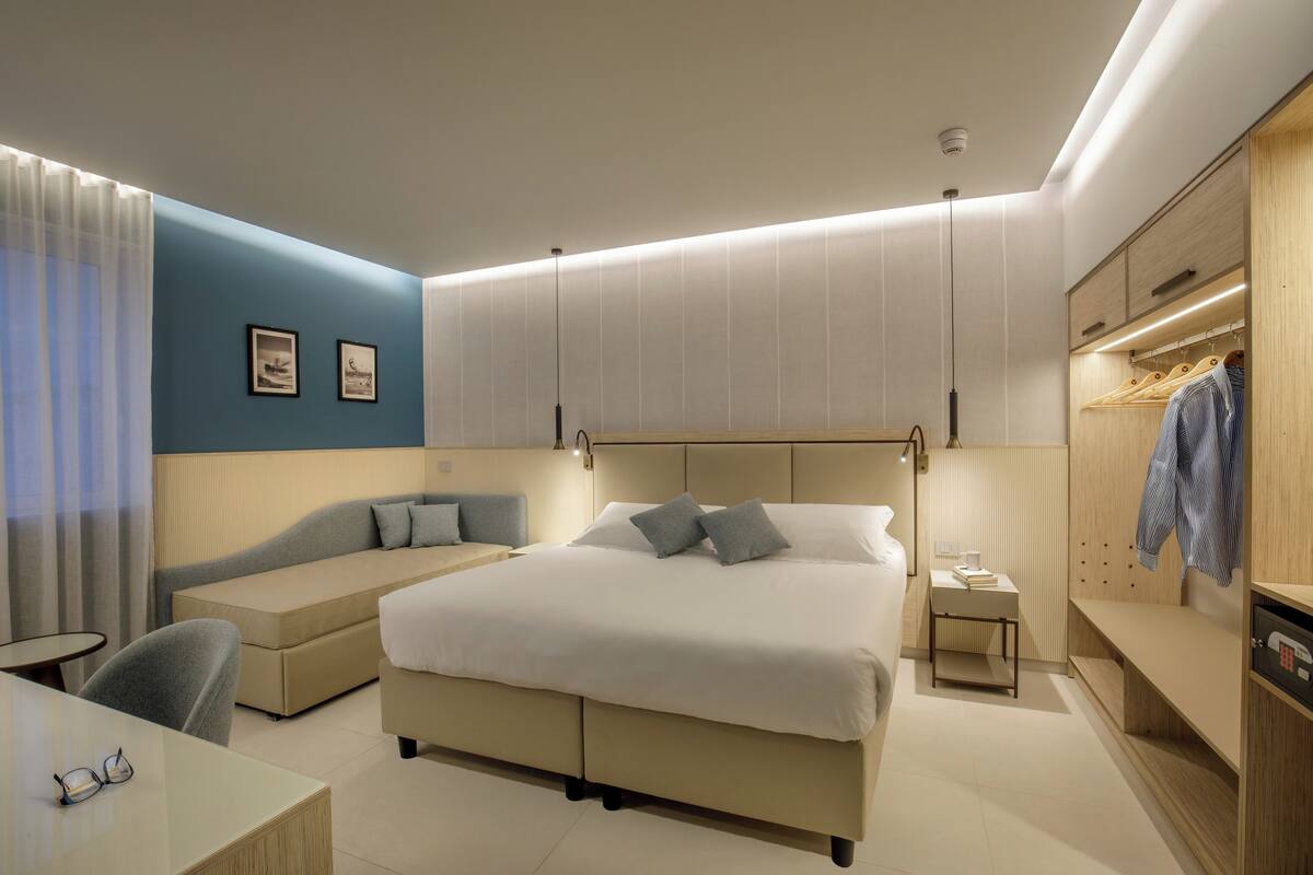 Quarto de hotel moderno com uma cama grande, sofá, escrivaninha e guarda-roupa.