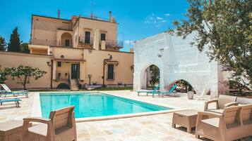 Uma piscina com espreguiçadeiras, uma estrutura branca com arcos e um edifício de vários andares ao fundo.