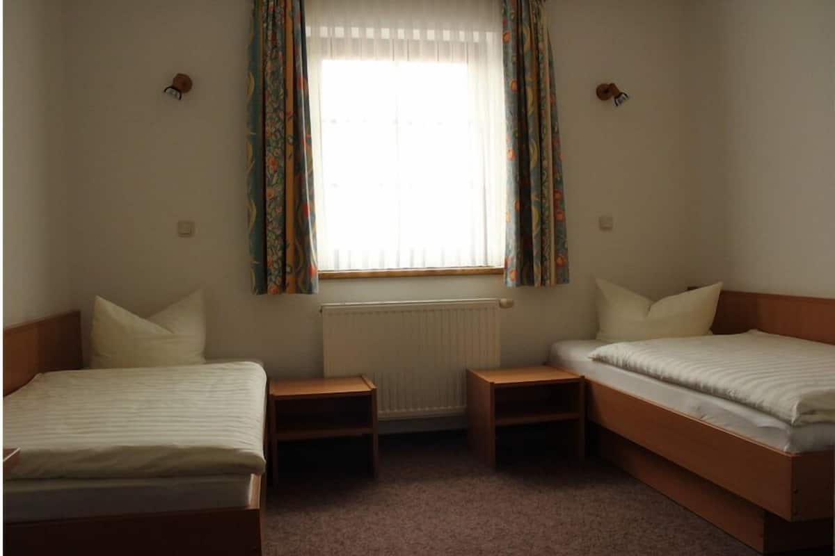 Quarto com duas camas individuais, janela com cortinas, radiador e duas luminárias fixadas na parede.