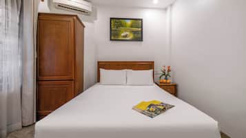 Cama bem arrumada com roupa de cama branca e uma revista, guarda-roupa de madeira, mesa de cabeceira com um vaso de flores e um quadro de uma paisagem na parede.