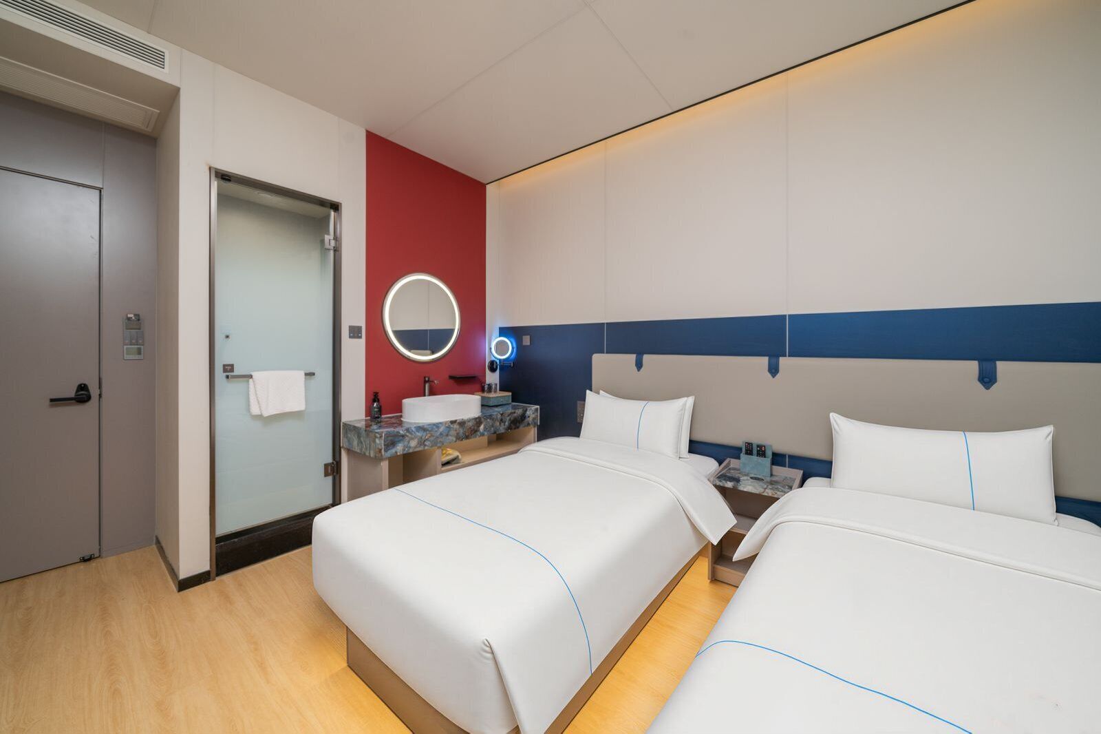 Quarto de hotel com duas camas, banheiro com toucador e uma parede de destaque vermelha.