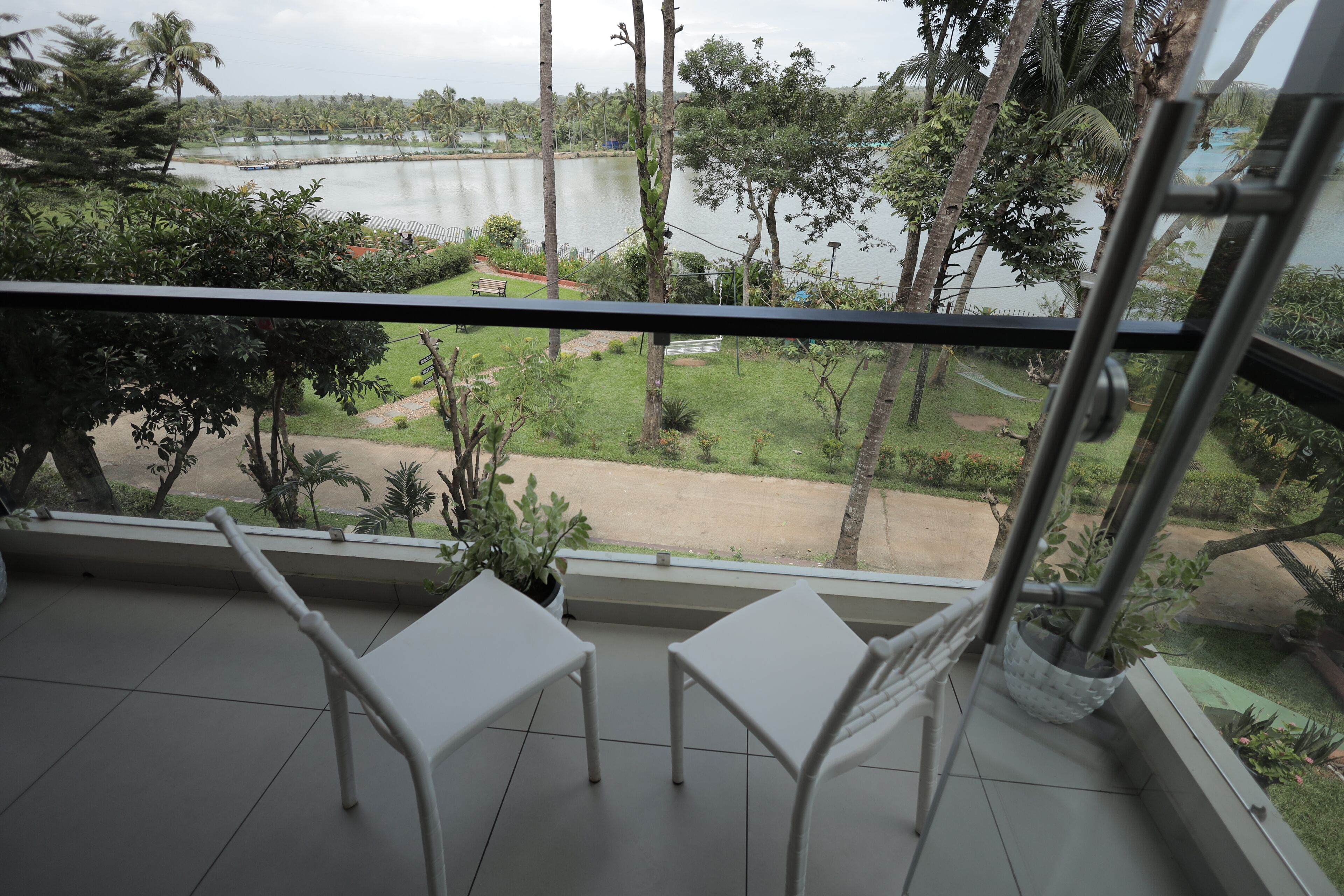 Varanda com duas cadeiras brancas, um vaso de plantas e vista para um lago com árvores e uma trilha.