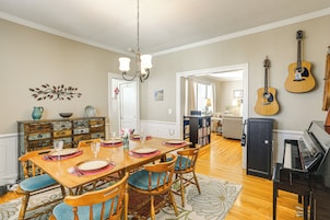 Uma sala de jantar com mesa de madeira posta para quatro pessoas, com cadeiras estofadas em azul de cada lado, um lustre acima e um piano à direita.