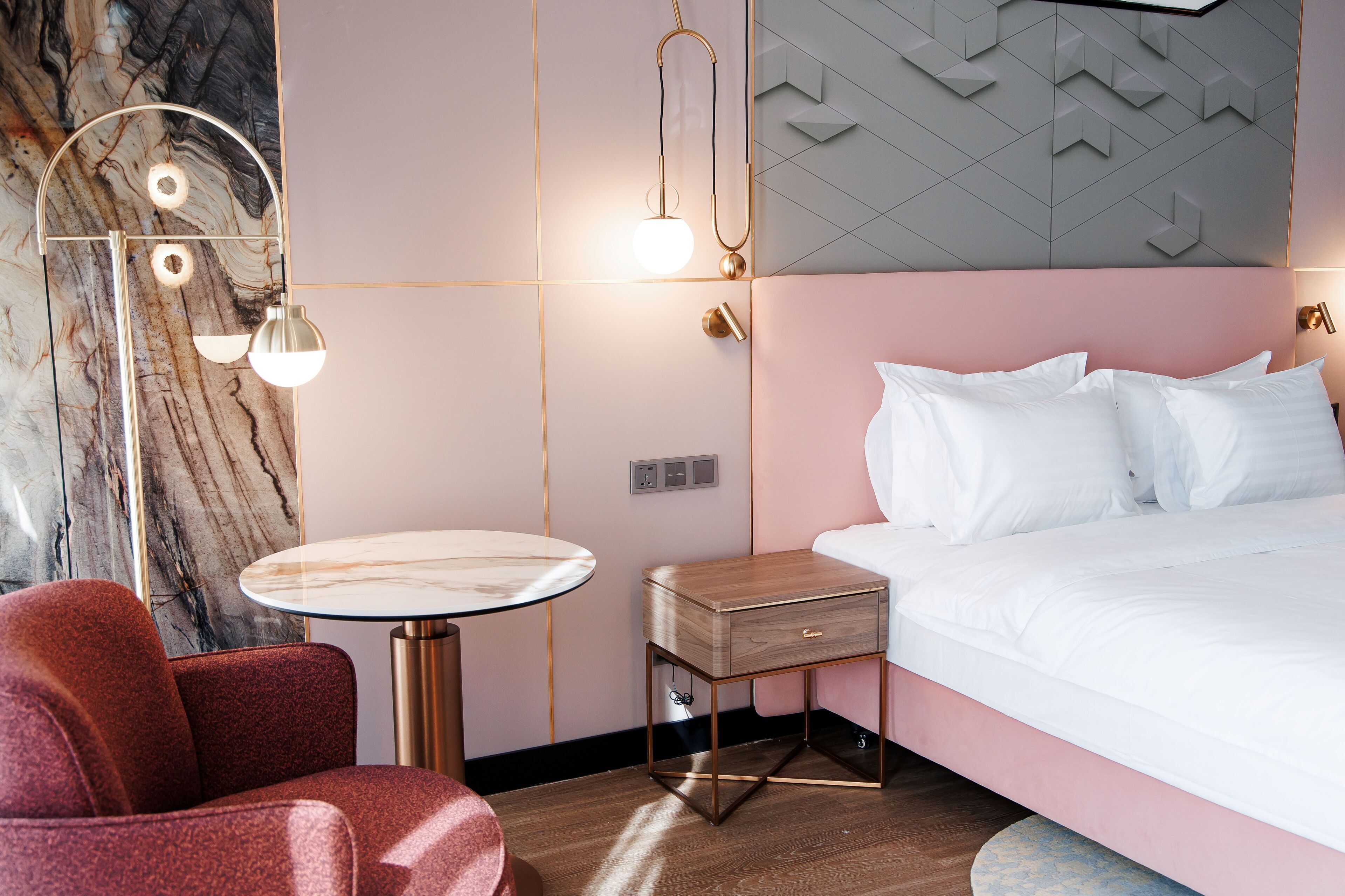 Quarto de hotel moderno com parede de mármore, uma mesa redonda, uma cadeira vermelha, uma mesinha de cabeceira e uma cama com roupa de cama branca.