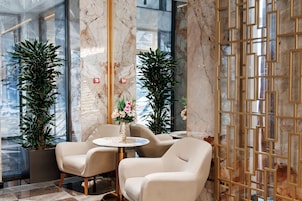 Um hall moderno com colunas de mármore, duas plantas em vasos, uma mesa redonda com cadeiras e um painel decorativo.