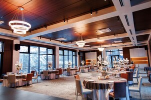 Uma ampla sala de banquetes com várias mesas preparadas para refeições, grandes janelas com vistas e luminárias modernas.