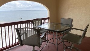 Varanda com mesa de vidro e quatro cadeiras, com vista para uma praia e o mar.