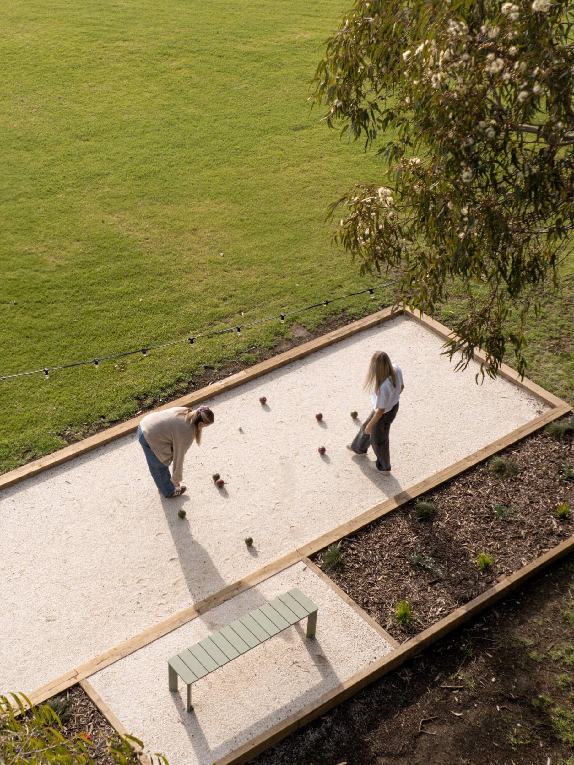 Uma pessoa jogando croquet em uma quadra de cascalho, com um banco e uma árvore ao fundo.