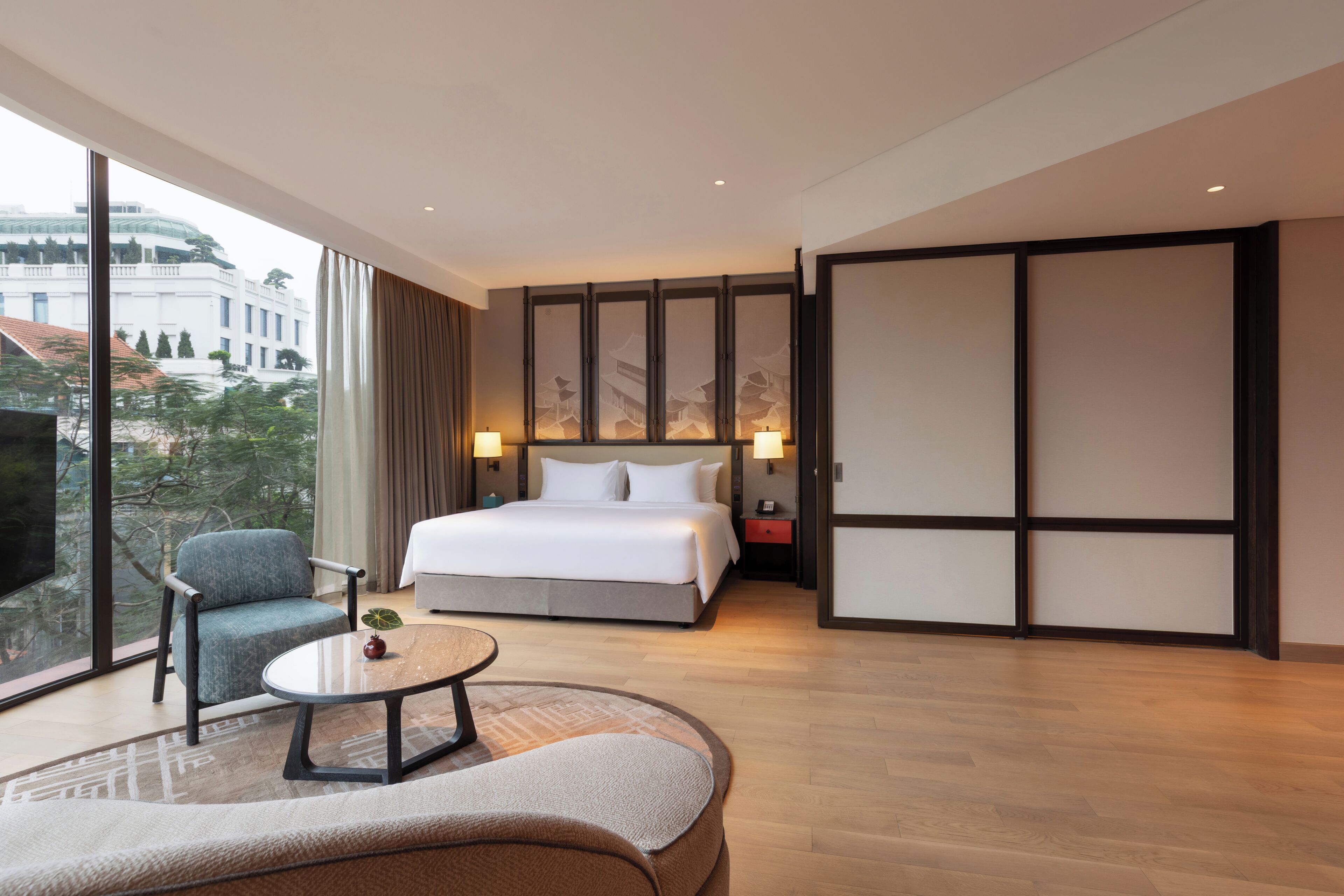 Quarto de hotel com uma cama grande, área de estar com mesa de centro, uma poltrona e vista para a cidade.