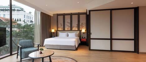 Quarto de hotel com uma cama grande, área de estar com mesa de centro, uma poltrona e vista para a cidade.