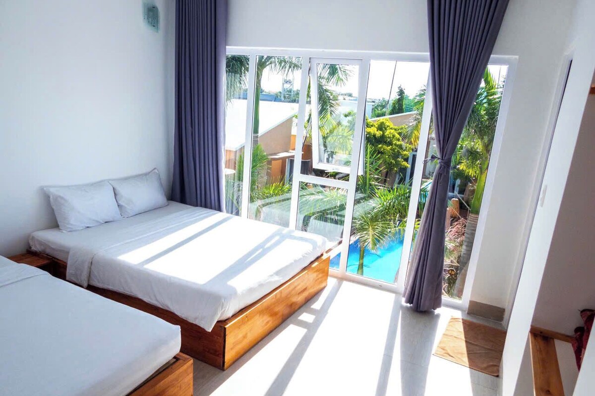 Quarto de hotel com uma grande porta de vidro deslizante que dá para uma área de piscina, duas camas e vista para palmeiras.
