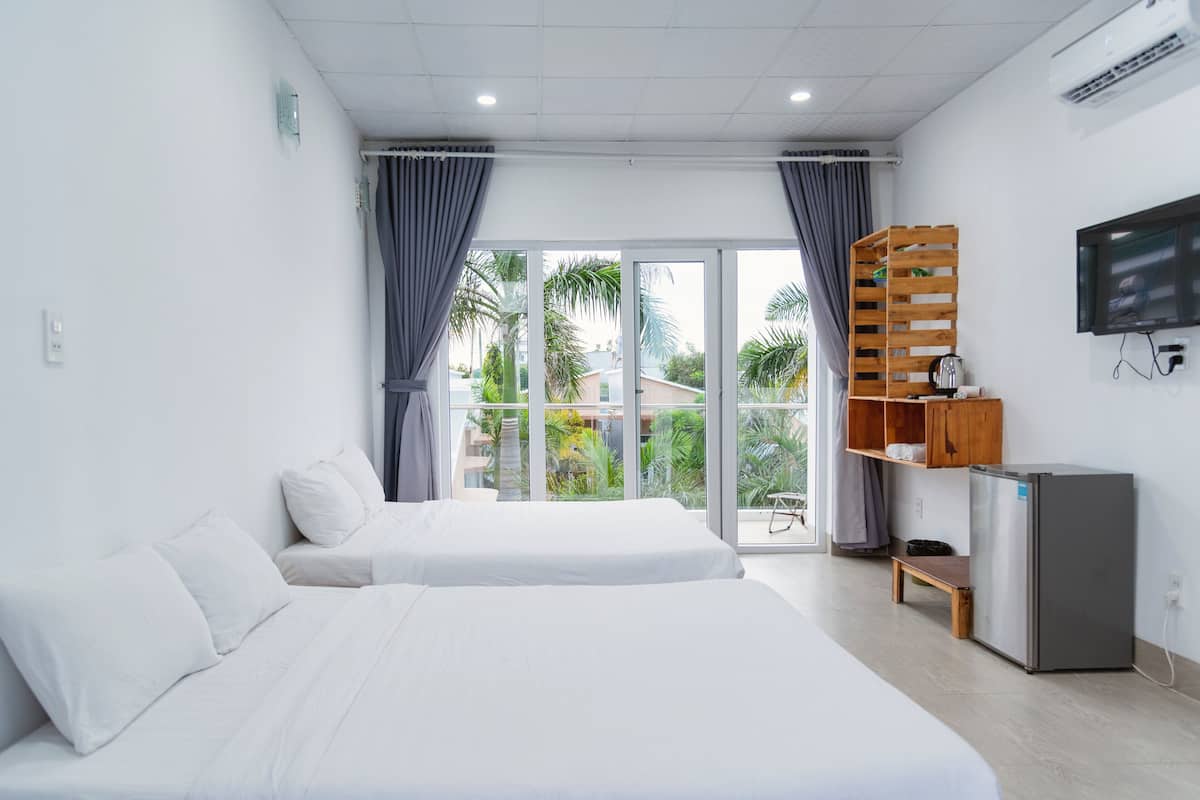 Quarto de hotel com duas camas, televisão, frigorífico e varanda com vista para palmeiras e edifícios.
