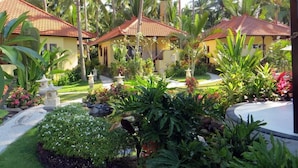 Um resort tropical com dois edifícios amarelos, palmeiras e um jardim bem cuidado com diversas plantas e flores.