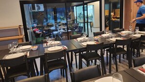 Uma sala de jantar com mesas postas para uma refeição, vista do interior do restaurante, e uma pessoa de camisa azul preparando comida.