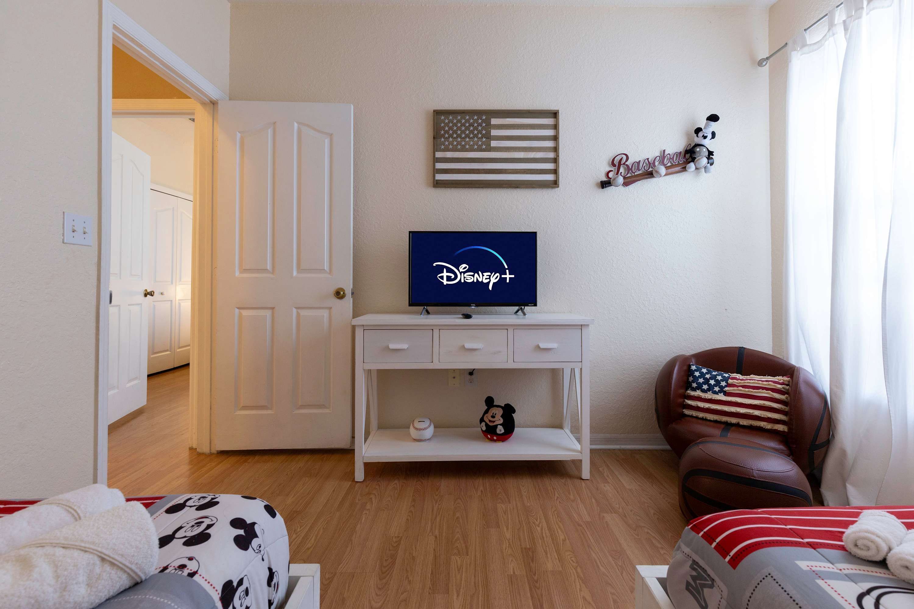 Quarto com televisão exibindo o logotipo do Disney+, uma escrivaninha com gavetas, uma cadeira com almofada decorada com a bandeira americana e uma prateleira com uma bola de beisebol e uma miniatura do Mickey Mouse.