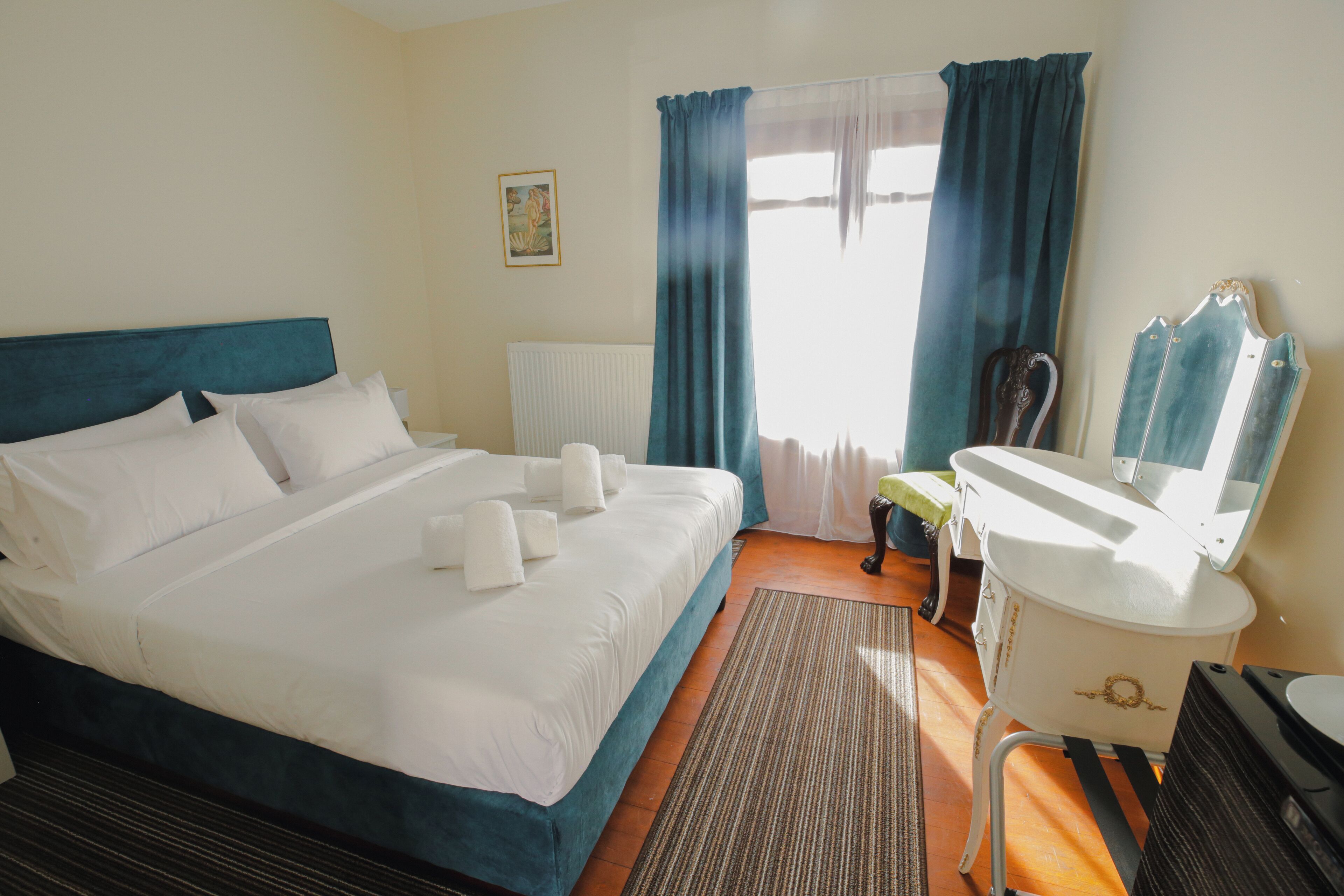 Quarto de hotel com cama, mesa de cabeceira, espelho, radiador e janela com cortinas.
