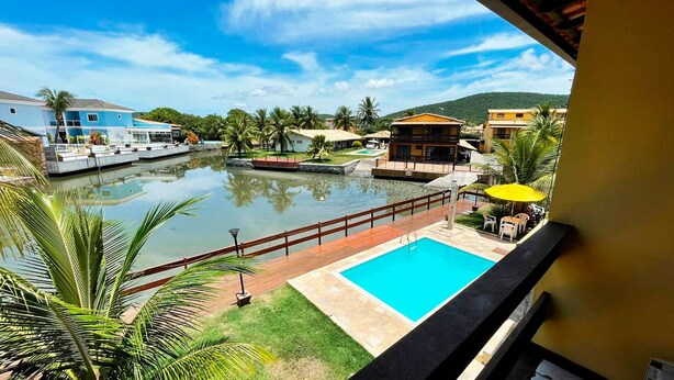 Uma área de piscina com deck de madeira, uma piscina pequena e vista para um corpo d'água e edifícios ao fundo.