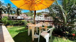 Um pátio com mesa e cadeiras sob um guarda-sol amarelo, cercado por vegetação exuberante e palmeiras.