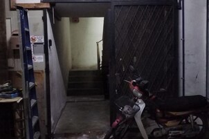 Um corredor estreito com escada que leva a um andar superior, uma motocicleta encostada na parede e um portão de metal.