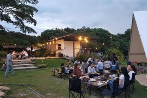 Reunião ao ar livre com pessoas sentadas em mesas, uma estrutura coberta e uma tenda em uma área gramada.