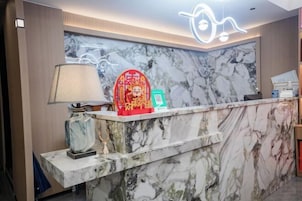 Balcão de recepção de hotel com bancada de mármore, uma luminária e um letreiro decorativo vermelho e verde.