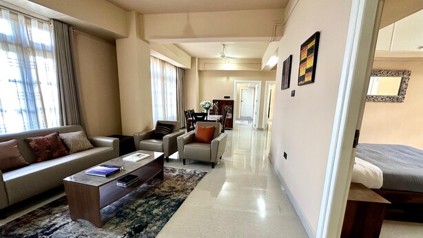 Uma sala de estar moderna com sofá, mesa de centro e cadeiras. Uma área de jantar é visível ao fundo, com uma mesa e cadeiras.