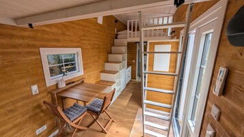 Quarto compacto com decoração em madeira, uma mesinha com cadeiras, uma janela e um mezanino acessado por uma escada.