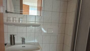 Casa de banho pequena e limpa, com lavatório branco, espelho e sanita. Zona de duche com porta de vidro.