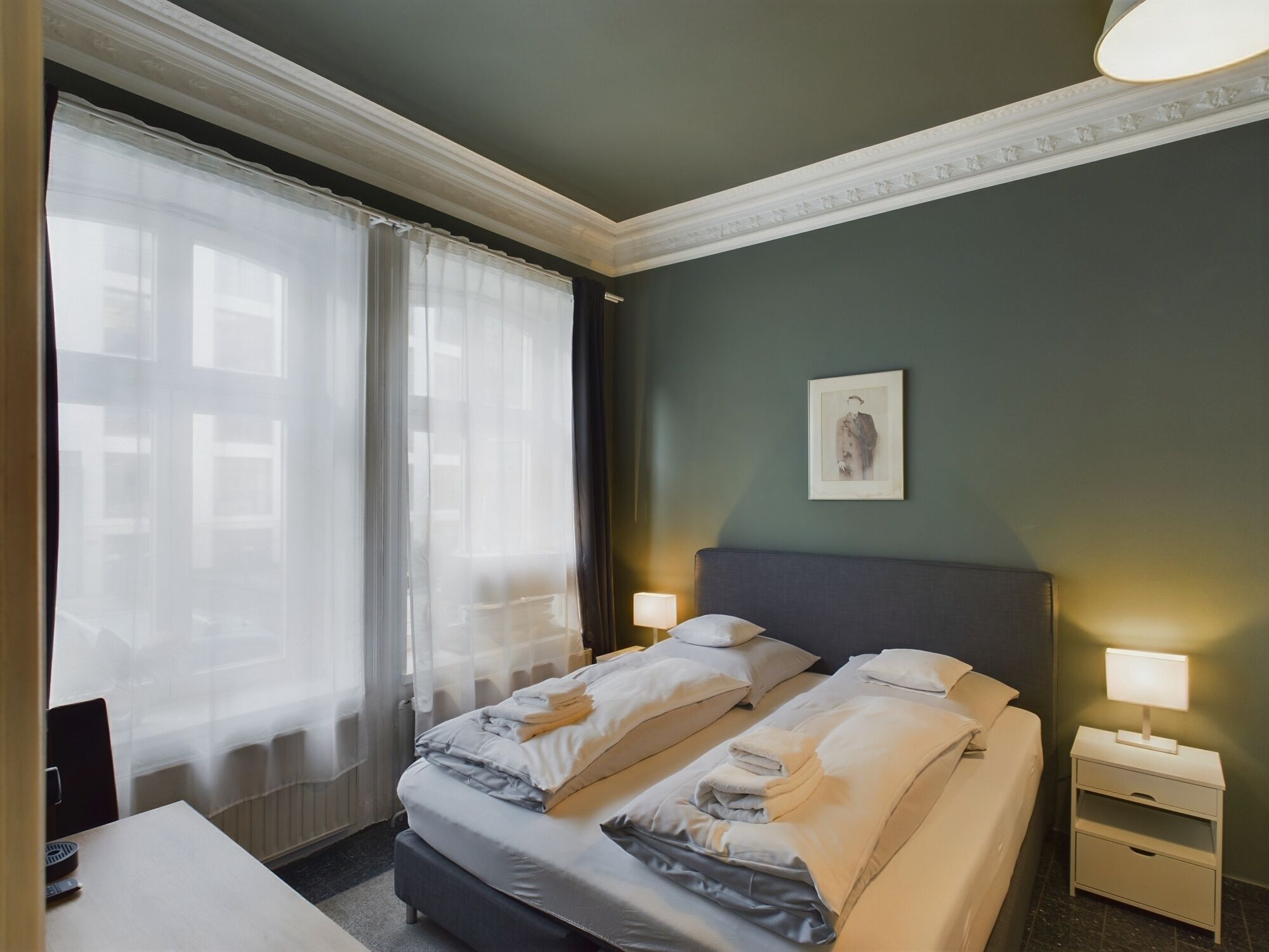 Quarto de hotel com uma cama, duas mesas de cabeceira, uma janela com cortinas translúcidas e um quadro na parede.