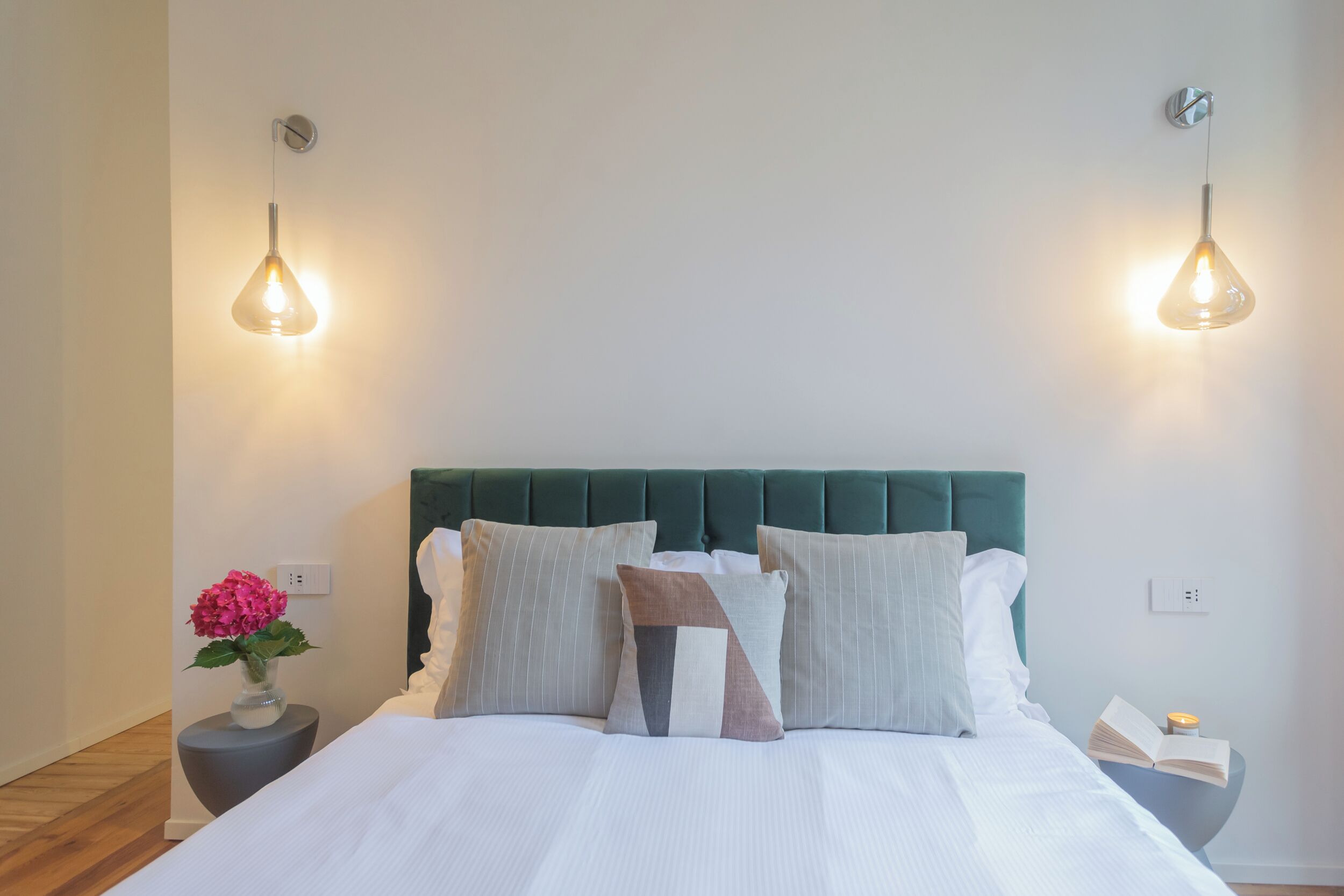 Cama bem arrumada com cabeceira estofada verde, duas mesas de cabeceira com abajures e um vaso com flores em uma das mesas.