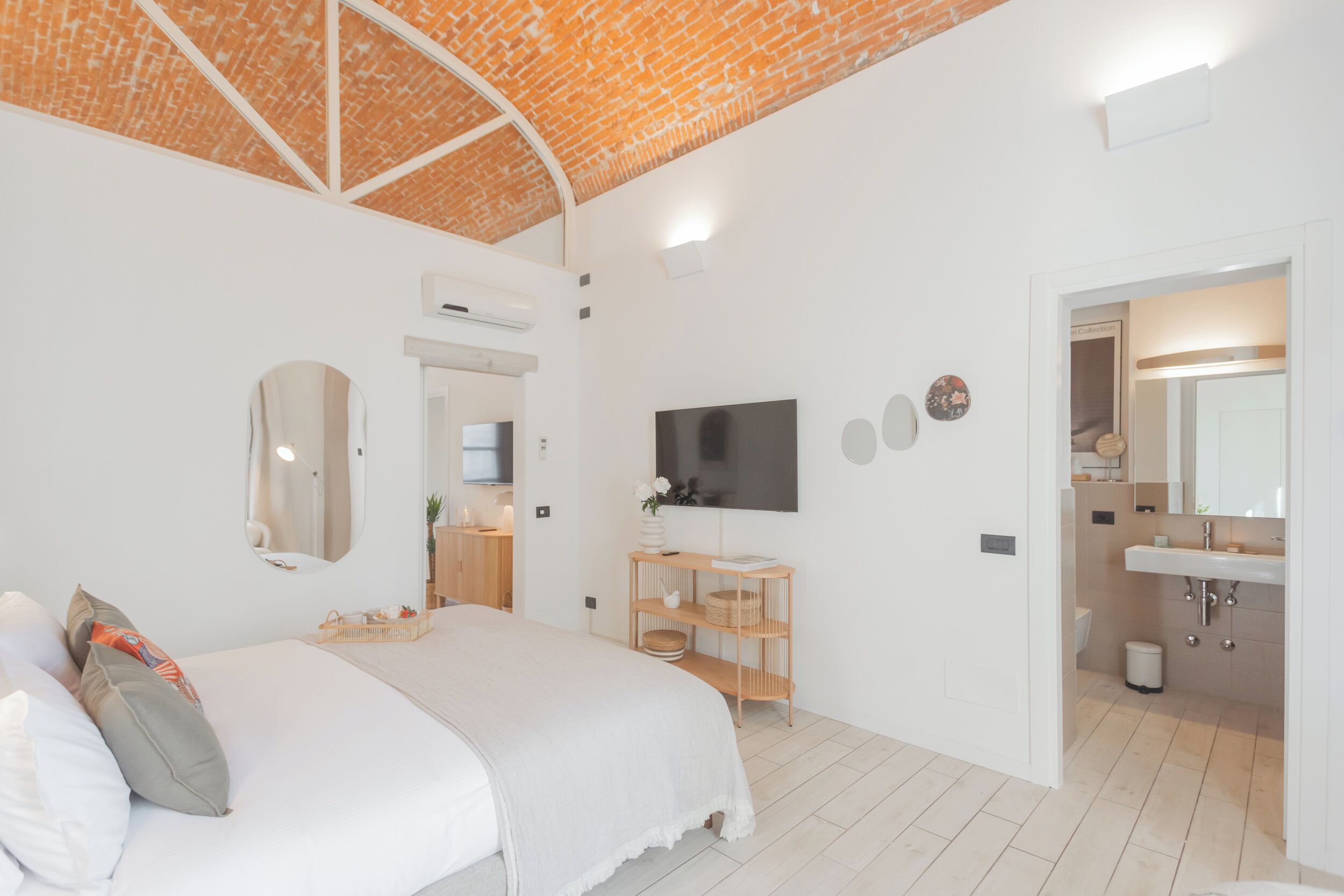 Quarto moderno com teto de tijolos, uma cama grande, uma TV e um banheiro com pia e espelho.