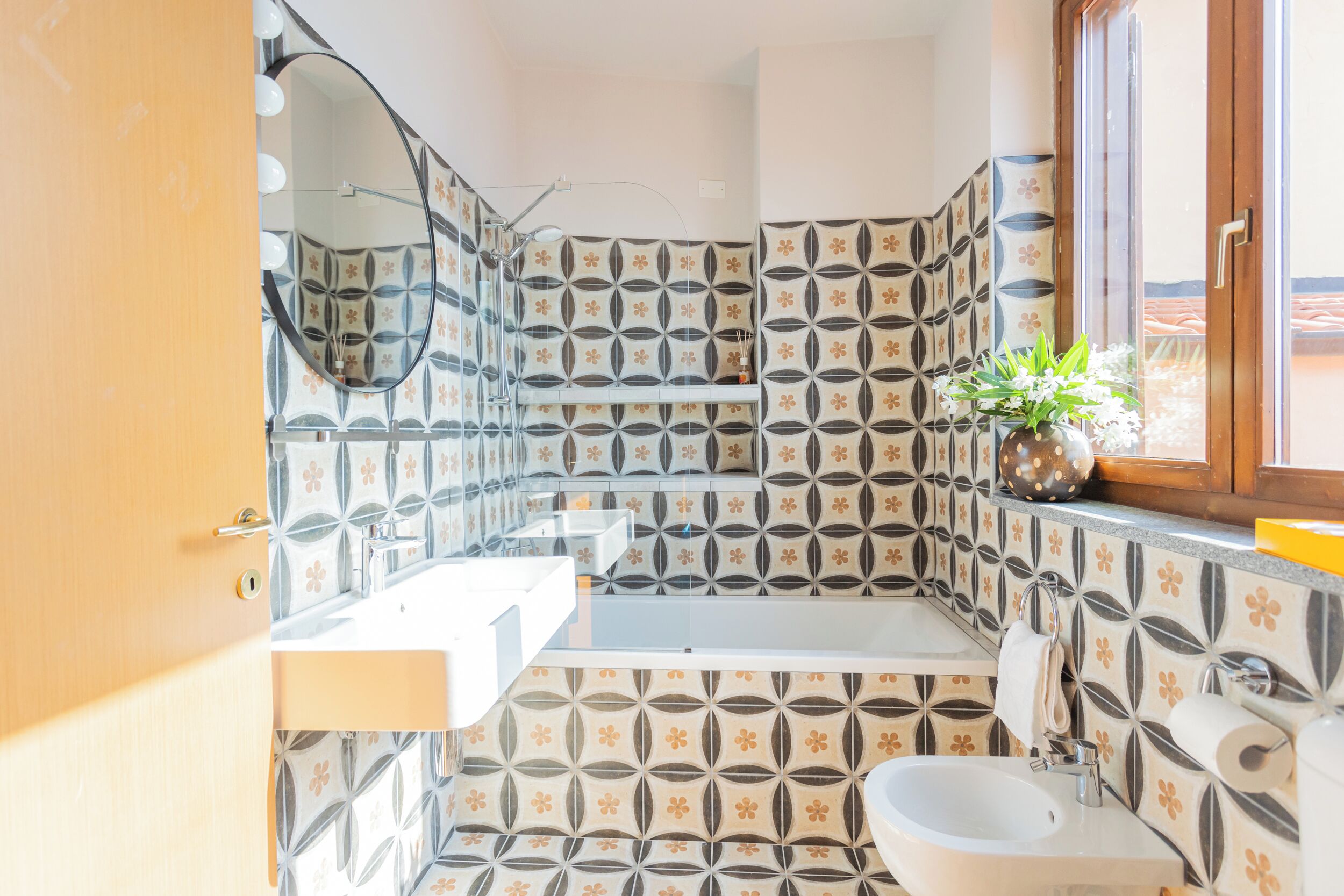 Um banheiro com azulejos estampados, um espelho, uma pia, um bidê e uma janela com vista.