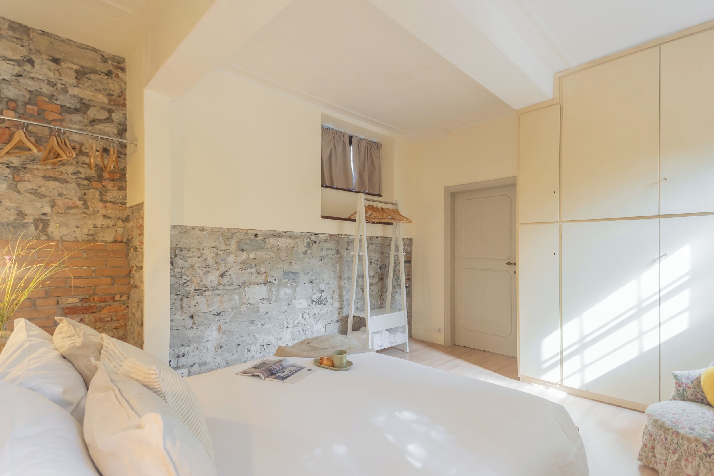 Quarto com cama, guarda-roupa, cabideiro e uma pequena mesa com uma chávena e um livro.