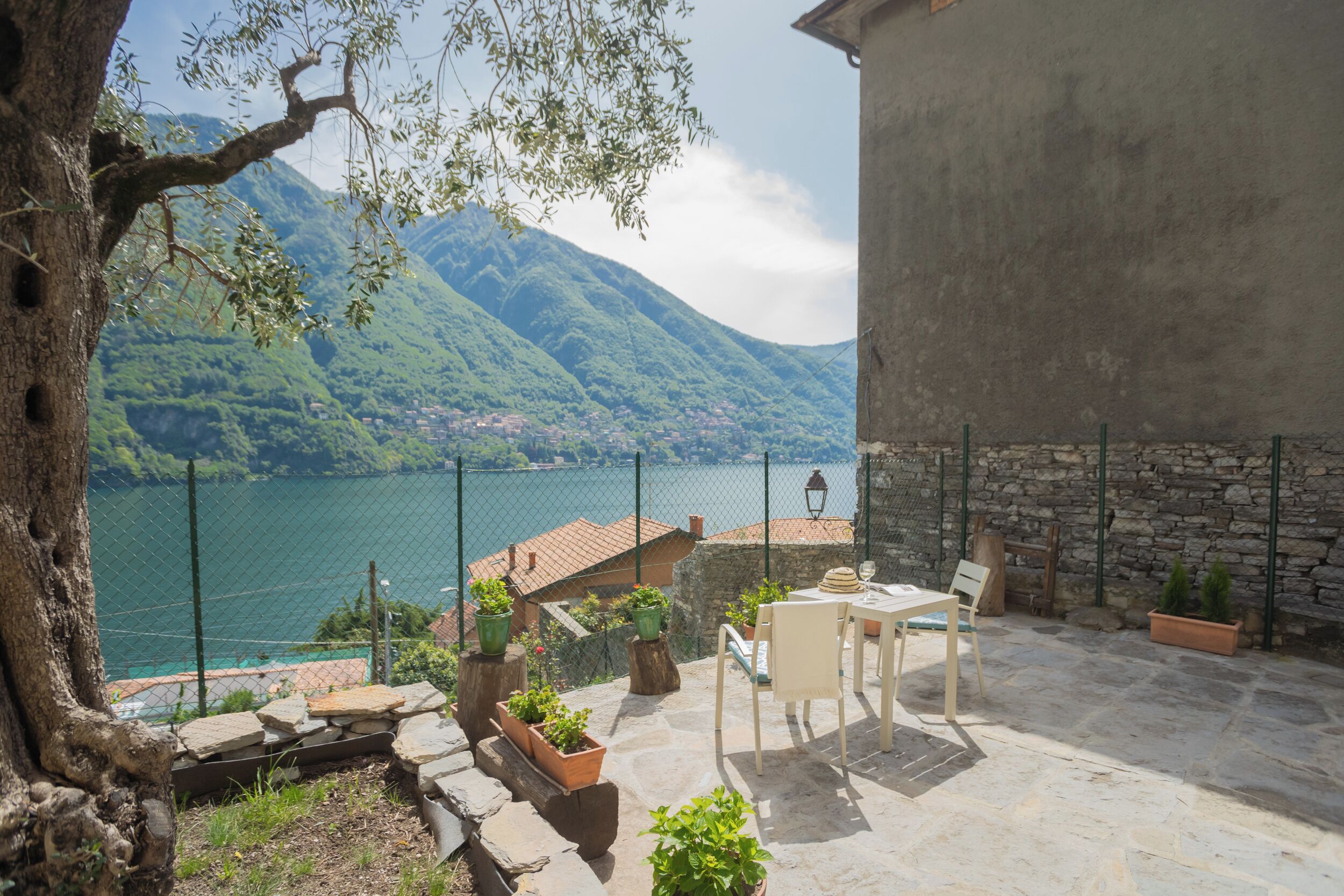 Um terraço com piso de pedra, uma mesa e cadeiras, plantas em vasos e vista para um lago e montanhas.