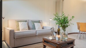 Uma sala de estar com um sofá, uma pequena mesa de madeira com uma vela, um vaso com uma planta e um radiador.