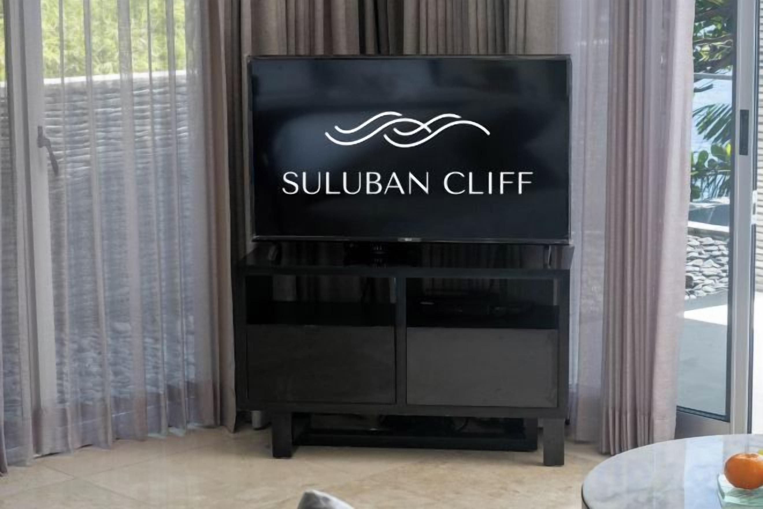 Uma unidade de entretenimento preta com uma TV de tela plana exibindo 'SULUBAN CLIFF' e uma pequena mesa com tampo de vidro e uma fruta laranja.