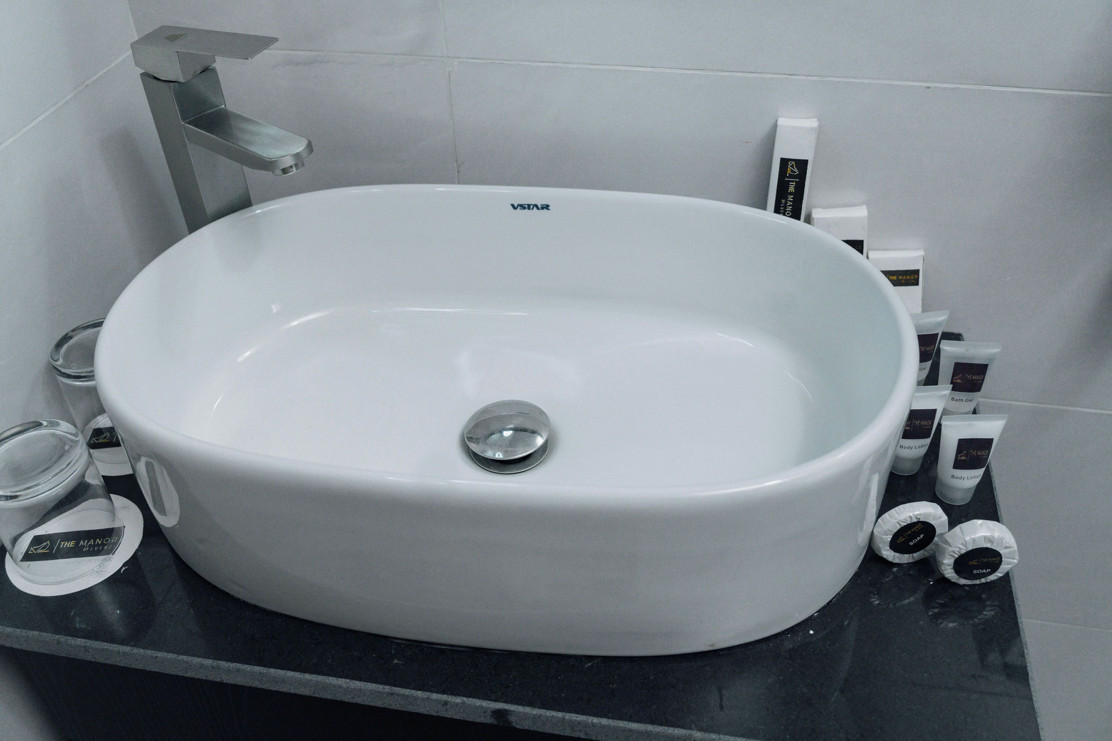 Uma pia branca oval com uma torneira moderna, rodeada por produtos de higiene e um bancada preta.