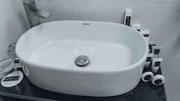 Uma pia branca oval com uma torneira moderna, rodeada por produtos de higiene e um bancada preta.