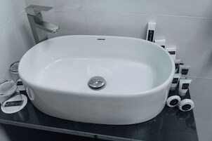 Uma pia branca oval com uma torneira moderna, rodeada por produtos de higiene e um bancada preta.
