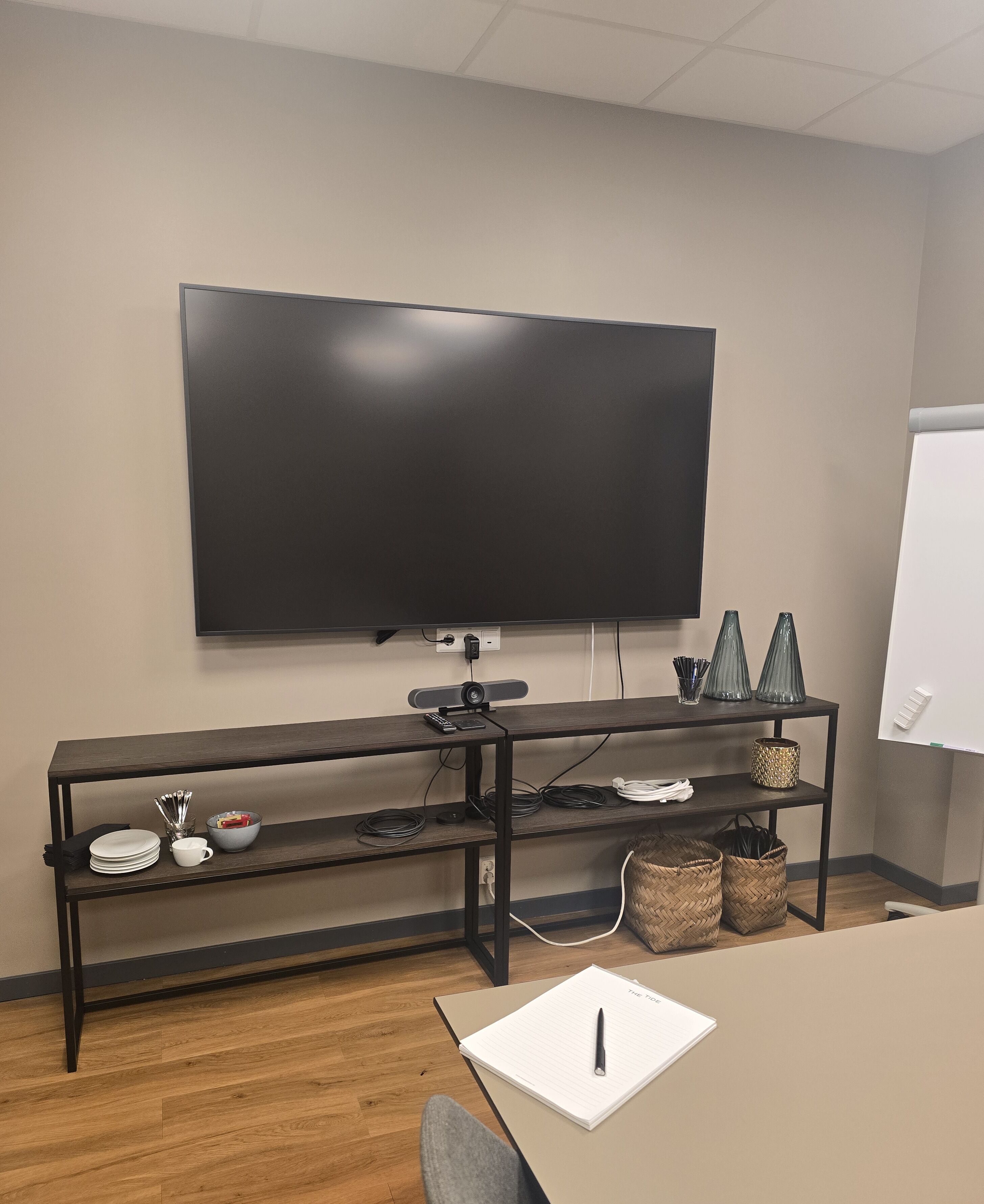 Uma sala de conferências com uma televisão de tela plana grande fixada na parede, uma mesa consola de madeira com diversos objetos e uma lousa branca à direita.