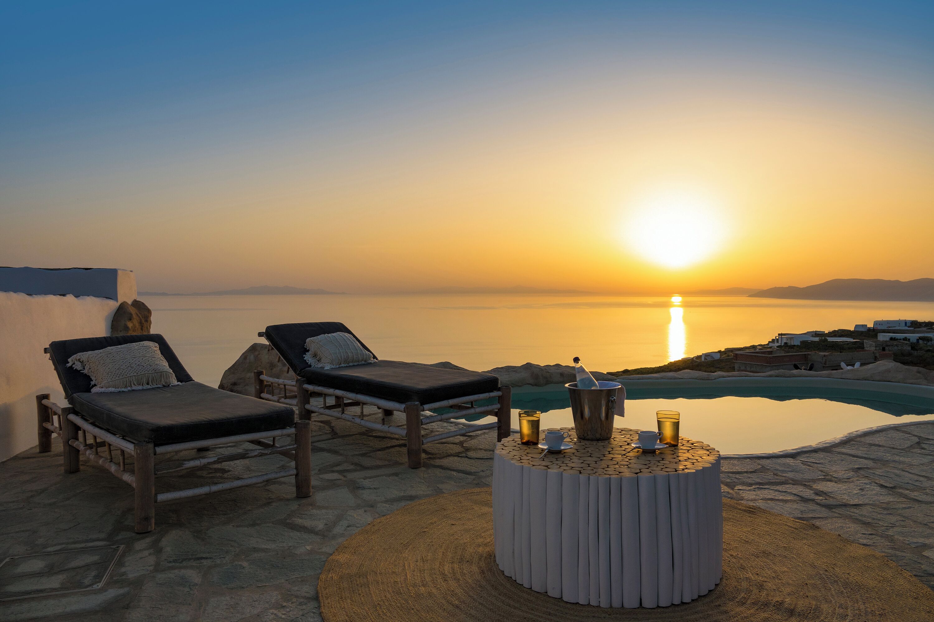 Vista do pôr do sol a partir de um terraço com duas cadeiras de descanso, uma mesa redonda com bebidas e uma piscina ao fundo.