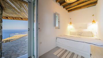 Quarto com soalho de pedra, cama com roupa de cama branca, teto de madeira e vista para o mar através de uma porta aberta.