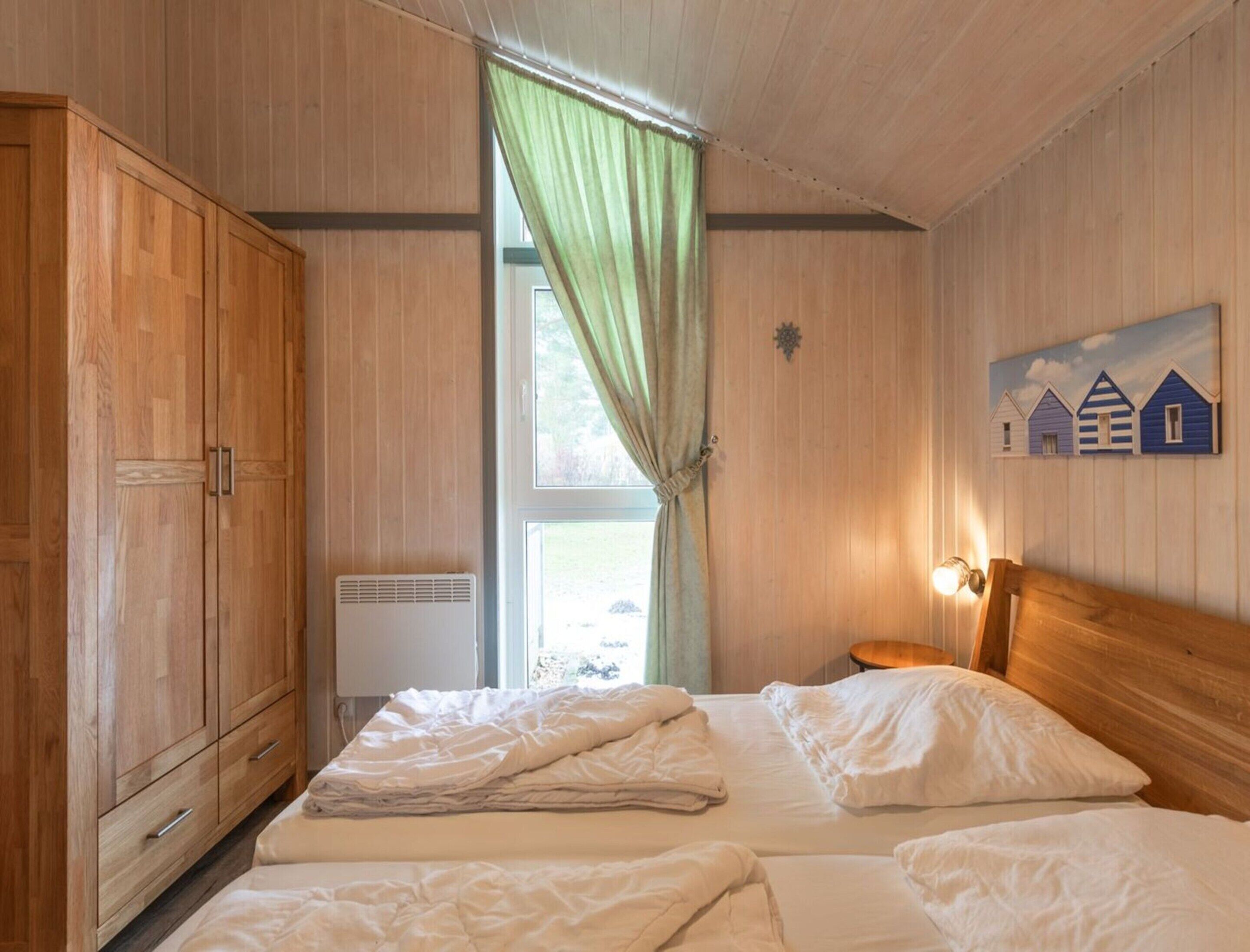 Quarto com duas camas, guarda-roupa de madeira, janela com cortinas verdes e um quadro na parede retratando casas.