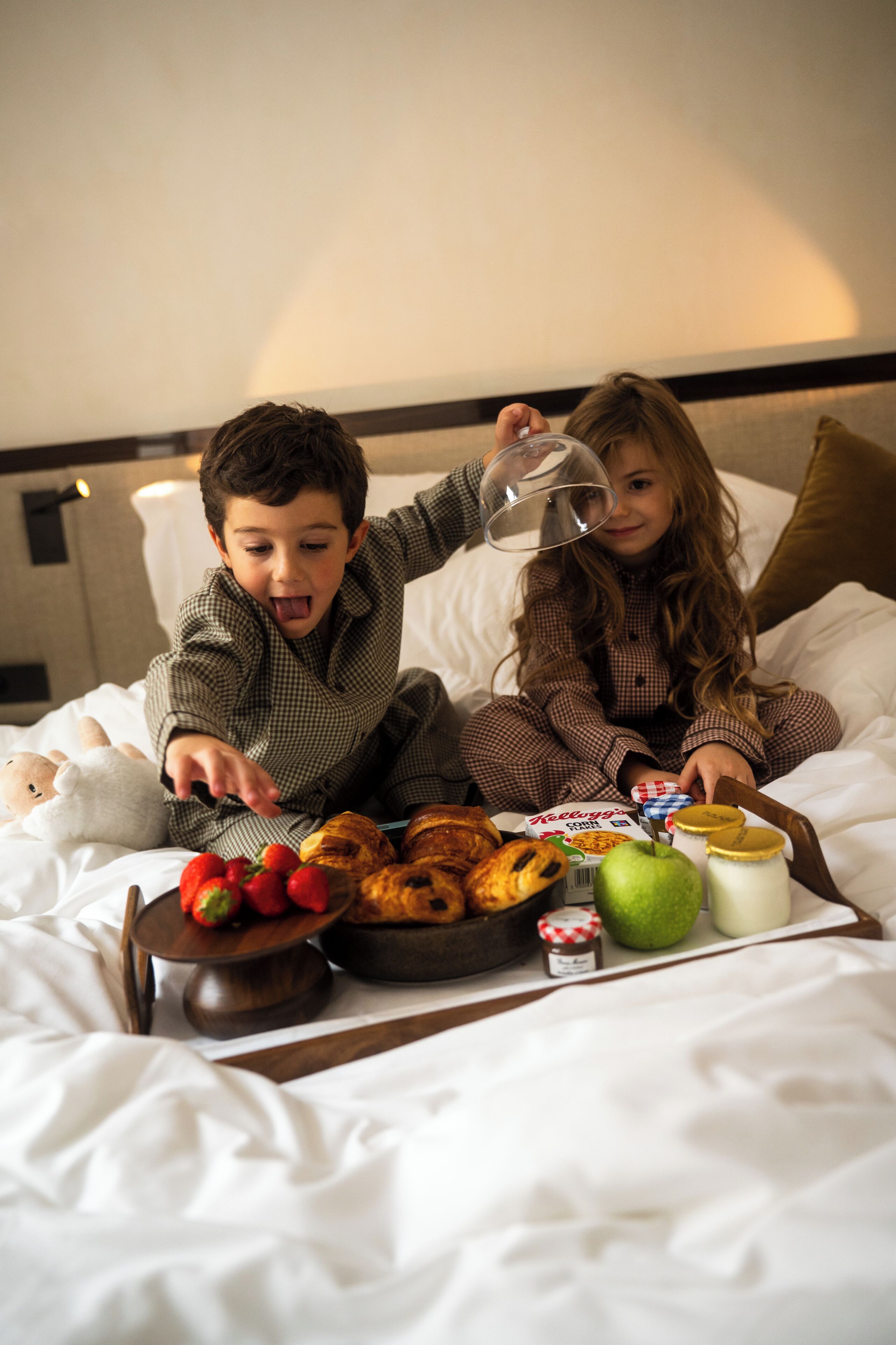 Duas crianças sentadas na cama com itens de café da manhã, incluindo morangos, croissants e cereais.